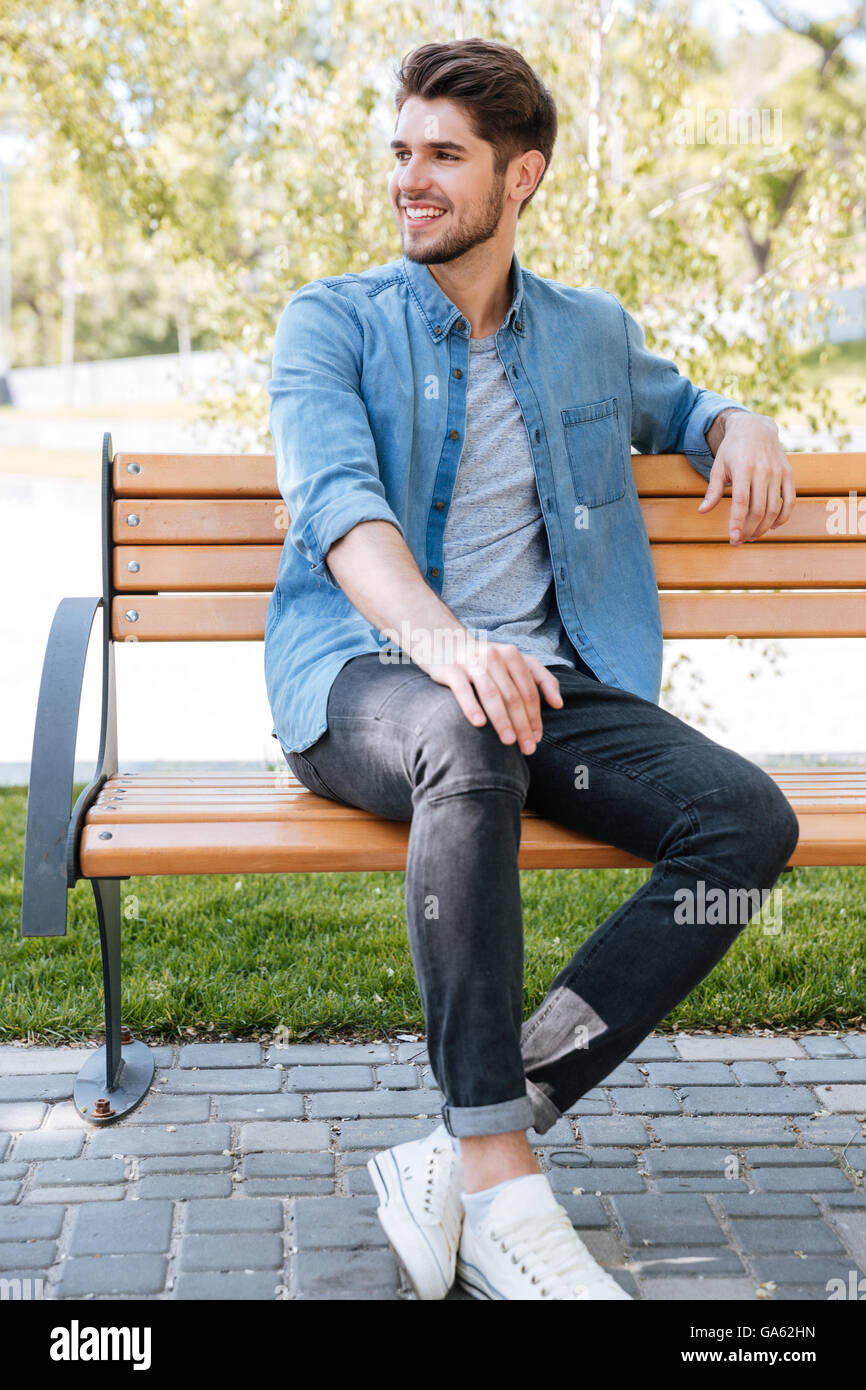 Handsome young man sitting on a bench smiling in the park Stock Photo ...