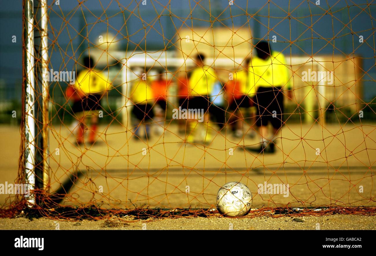 A football in the back of a goal net on a sand football pitch as two ...