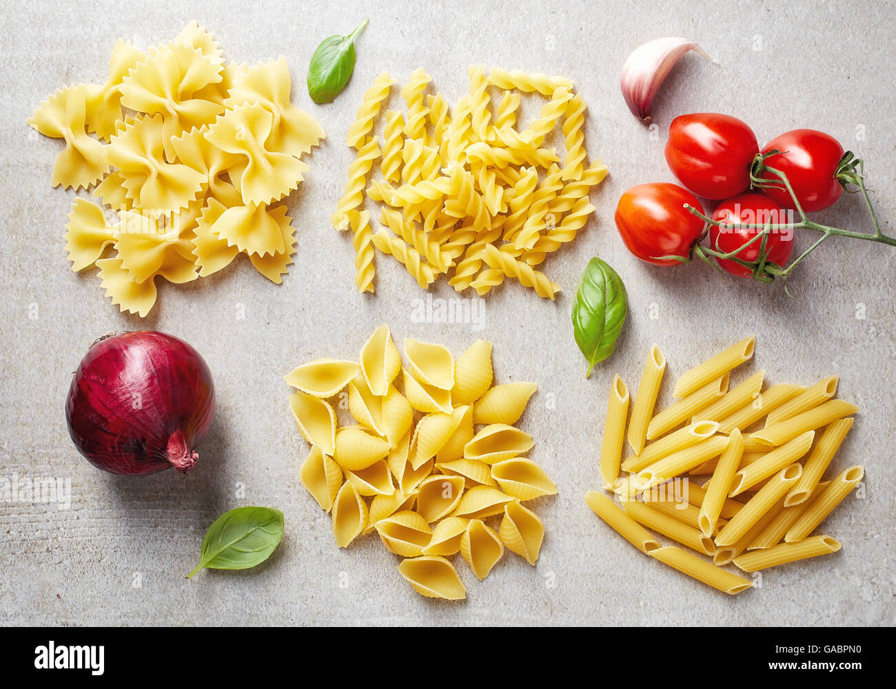 Various types of pasta, tomato, onion, garlic and basil leaves on grey ...
