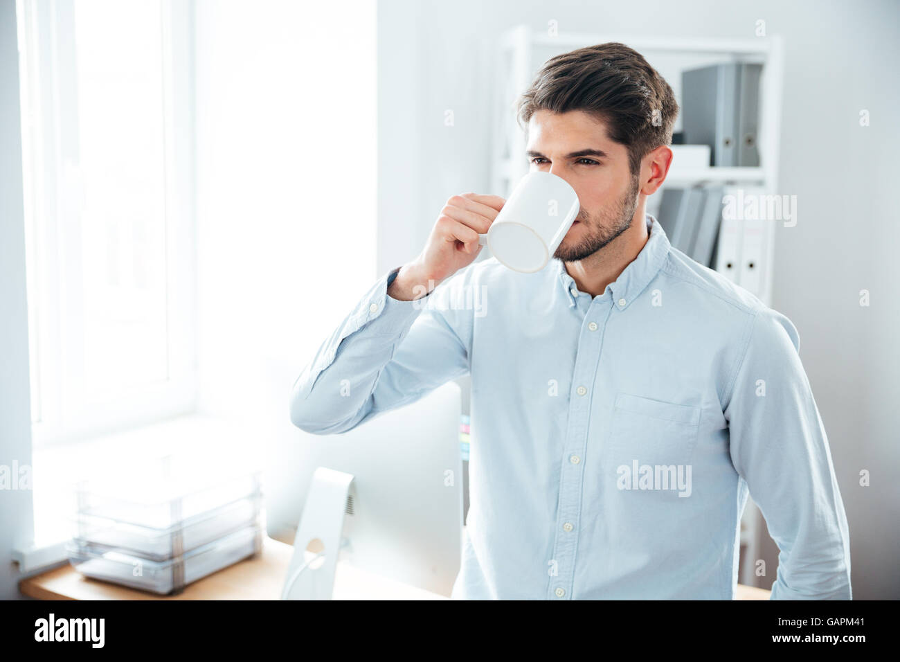 Handsome young man standing and drinking coffee in office Stock Photo ...