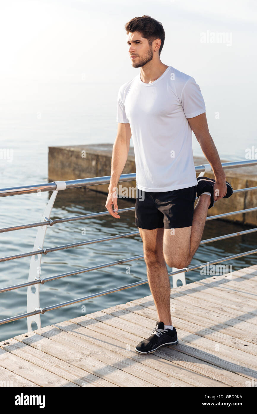 Attractive young sportsman doing exercises and stretching legs on pier ...