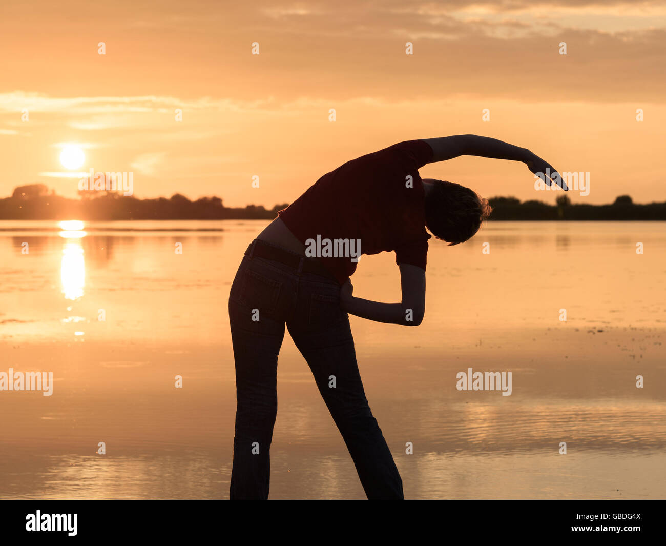 young slim woman doing exercises on the shore of the lake at sunset ...