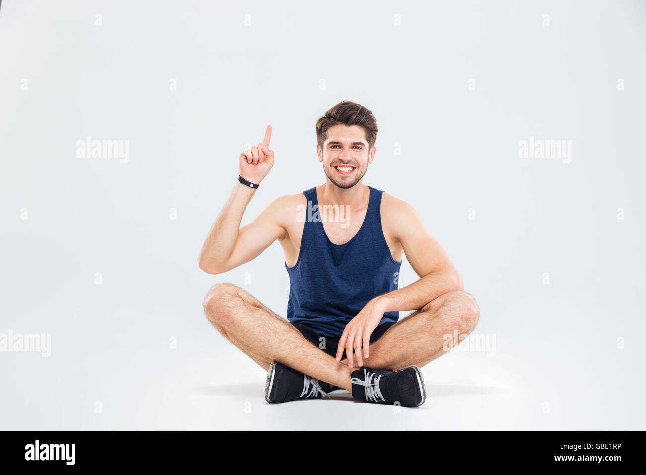 Cheerful young man athlete sitting with legs crossed and pointing up ...