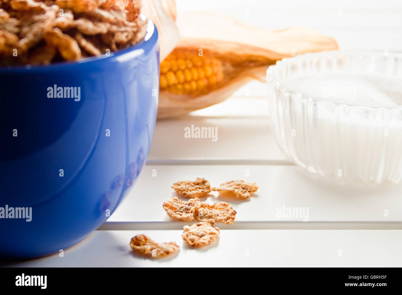 Cornflakes in a blue bowl on white wooden table with cob and sugar ...