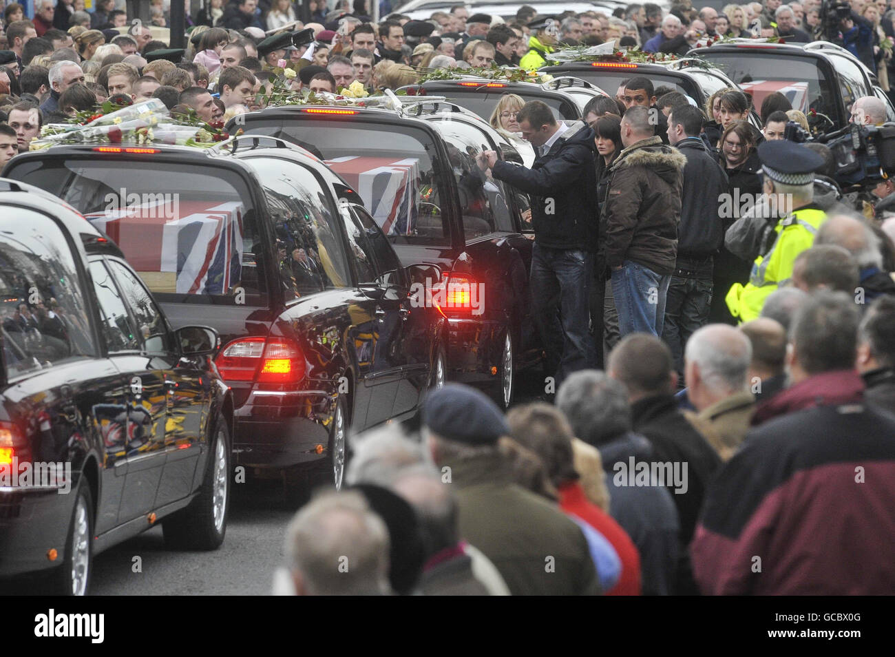 Mourners gather around the hearses and one man leans on the hearse ...