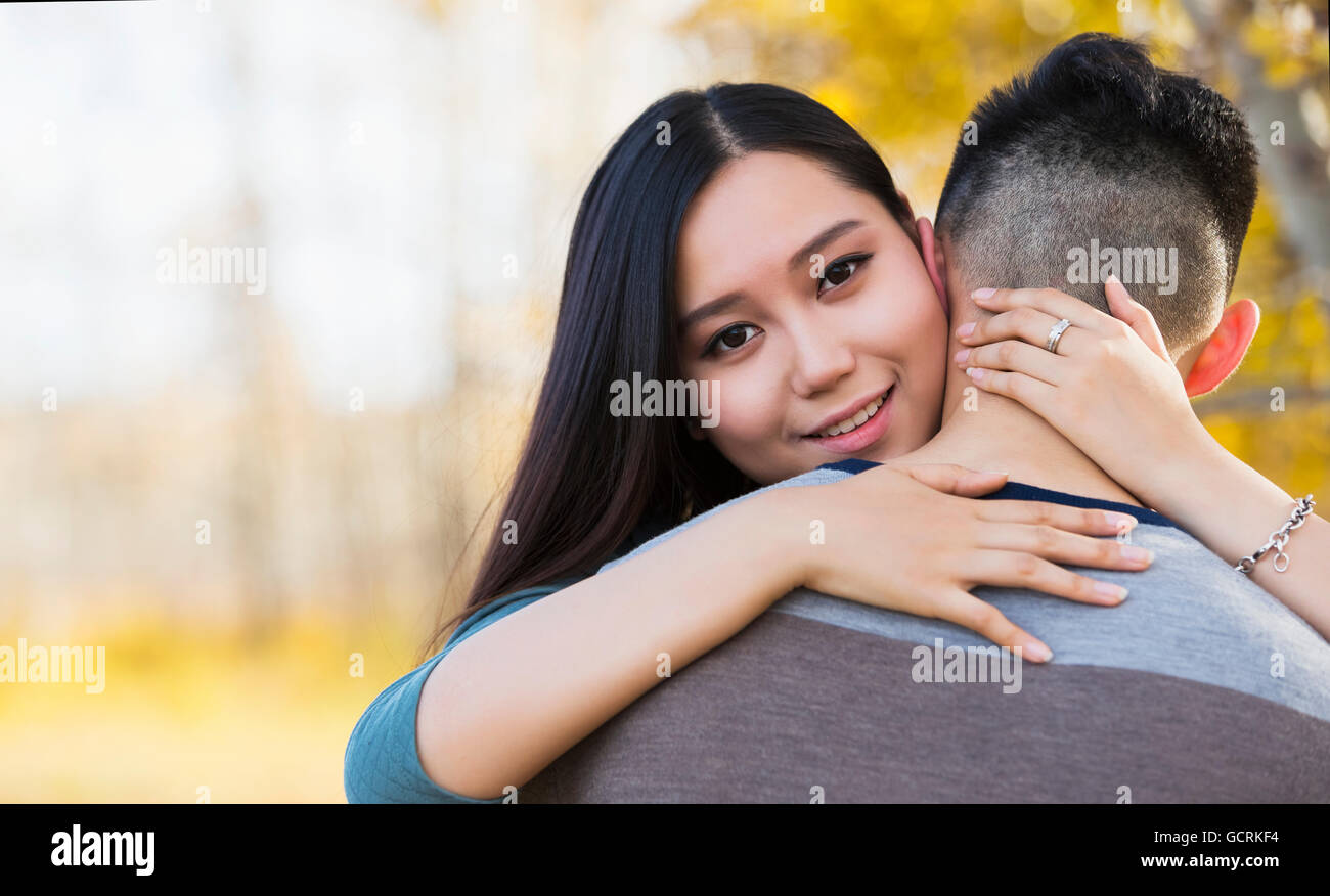 A young Asian woman hugging her boyfriend and looking over his shoulder at the camera in a park ...
