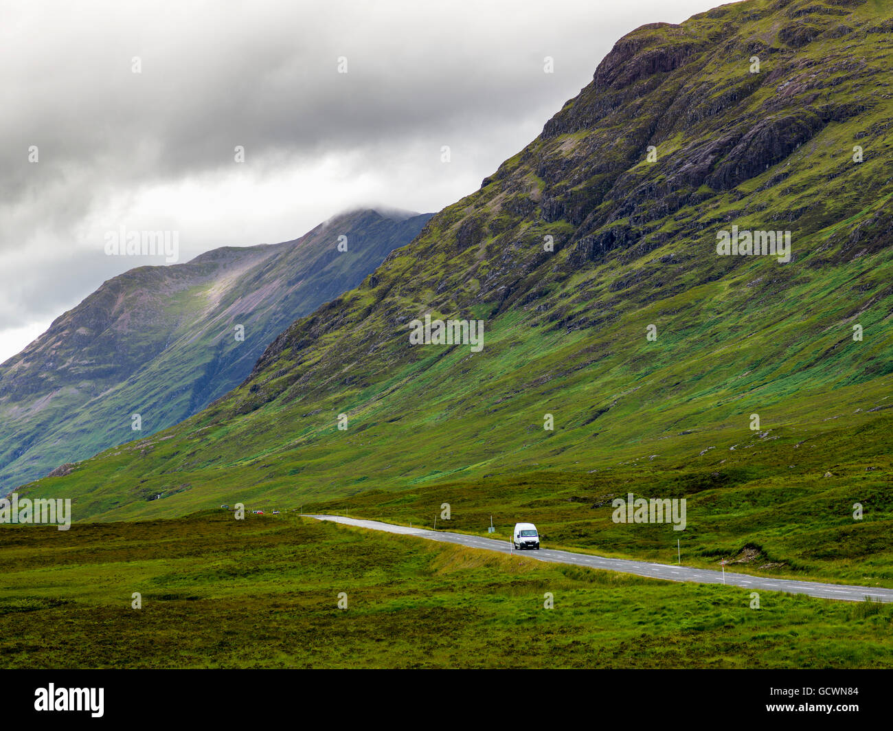 A vehicle travels on a road beside rugged mountains under a cloudy sky ...