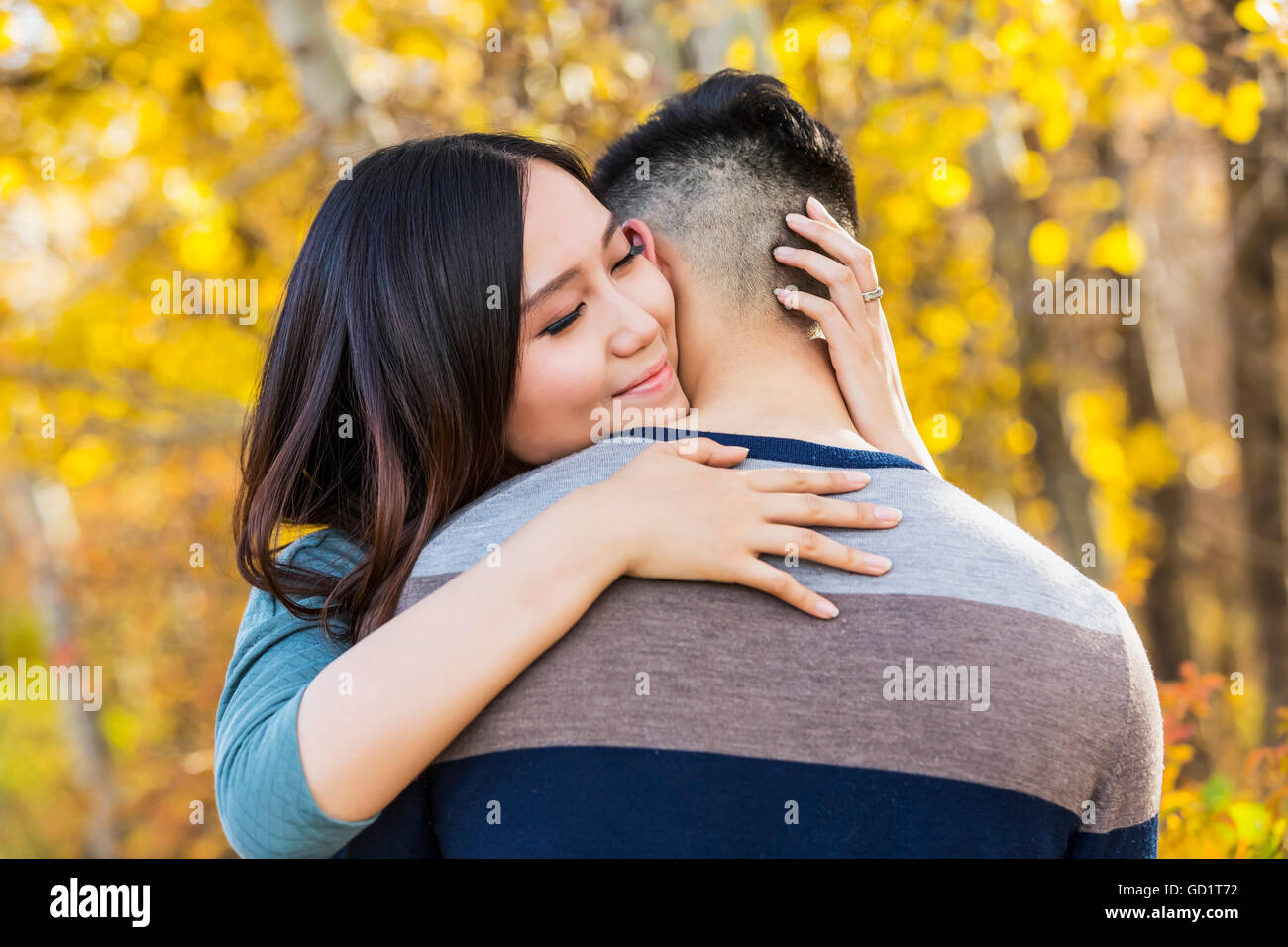 A young Asian woman hugging her boyfriend in a park in autumn and looking over his shoulder at ...