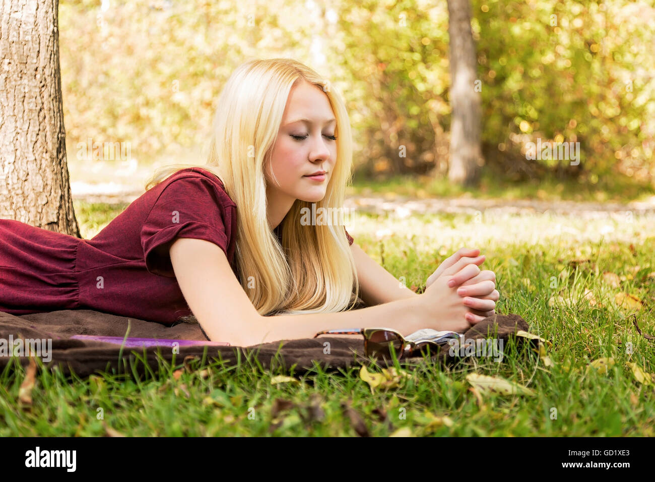 A young woman with long blond hair spending personal time in Bible study and praying outdoors in ...