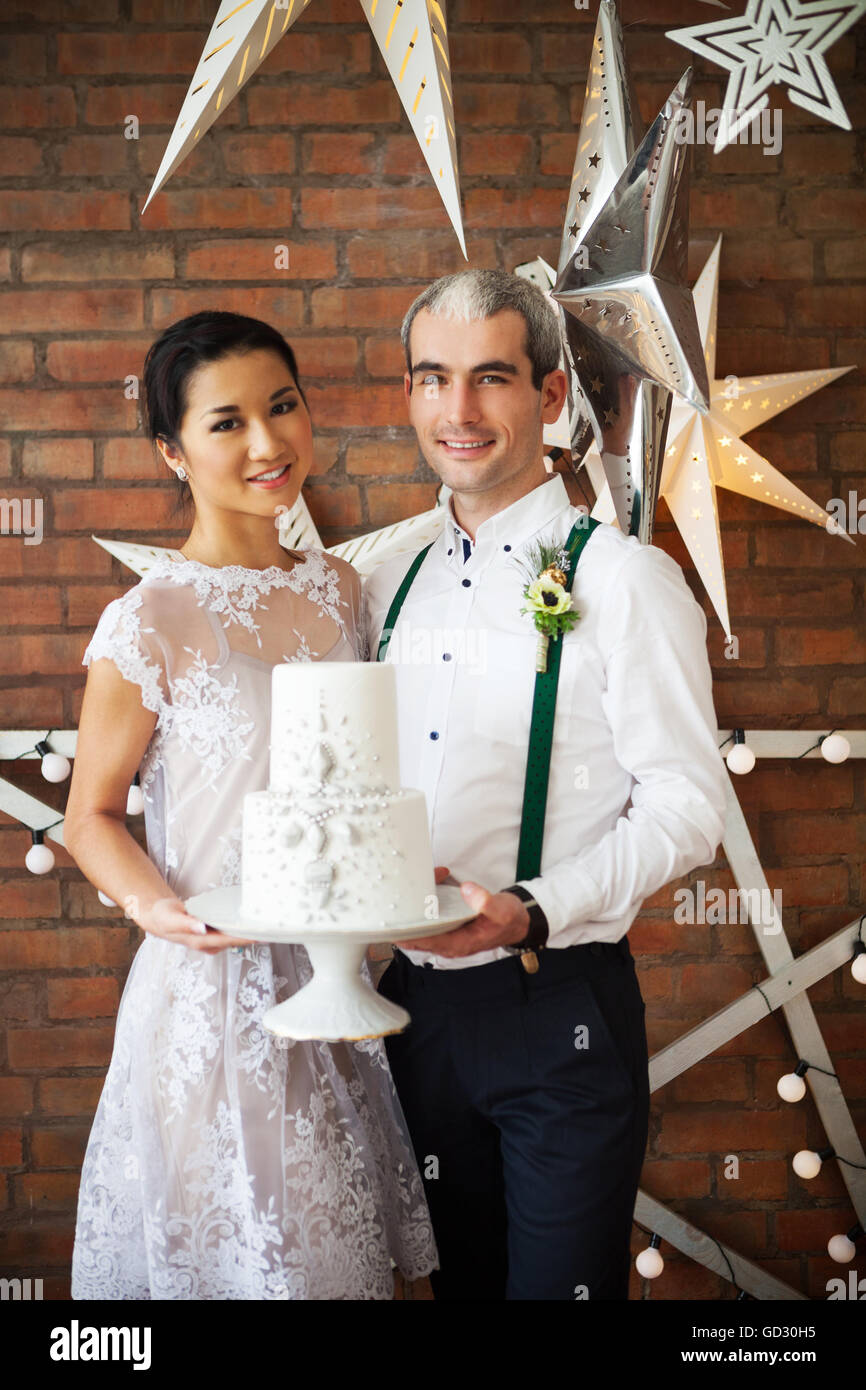 Cheerful married couple holding wedding cake near the brick wall ...