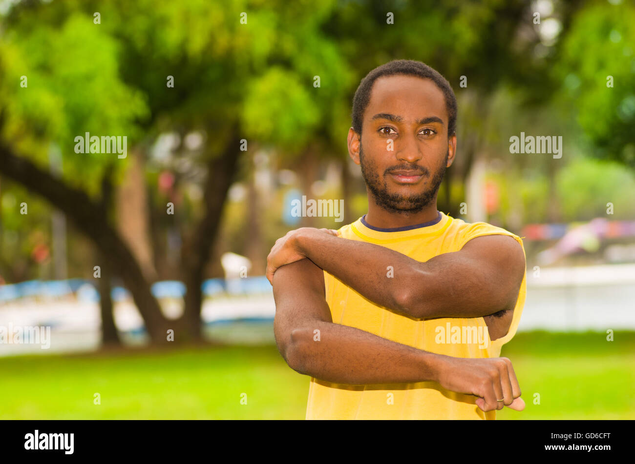 Man wearing yellow shirt stretching arms in park sorrounded by green ...