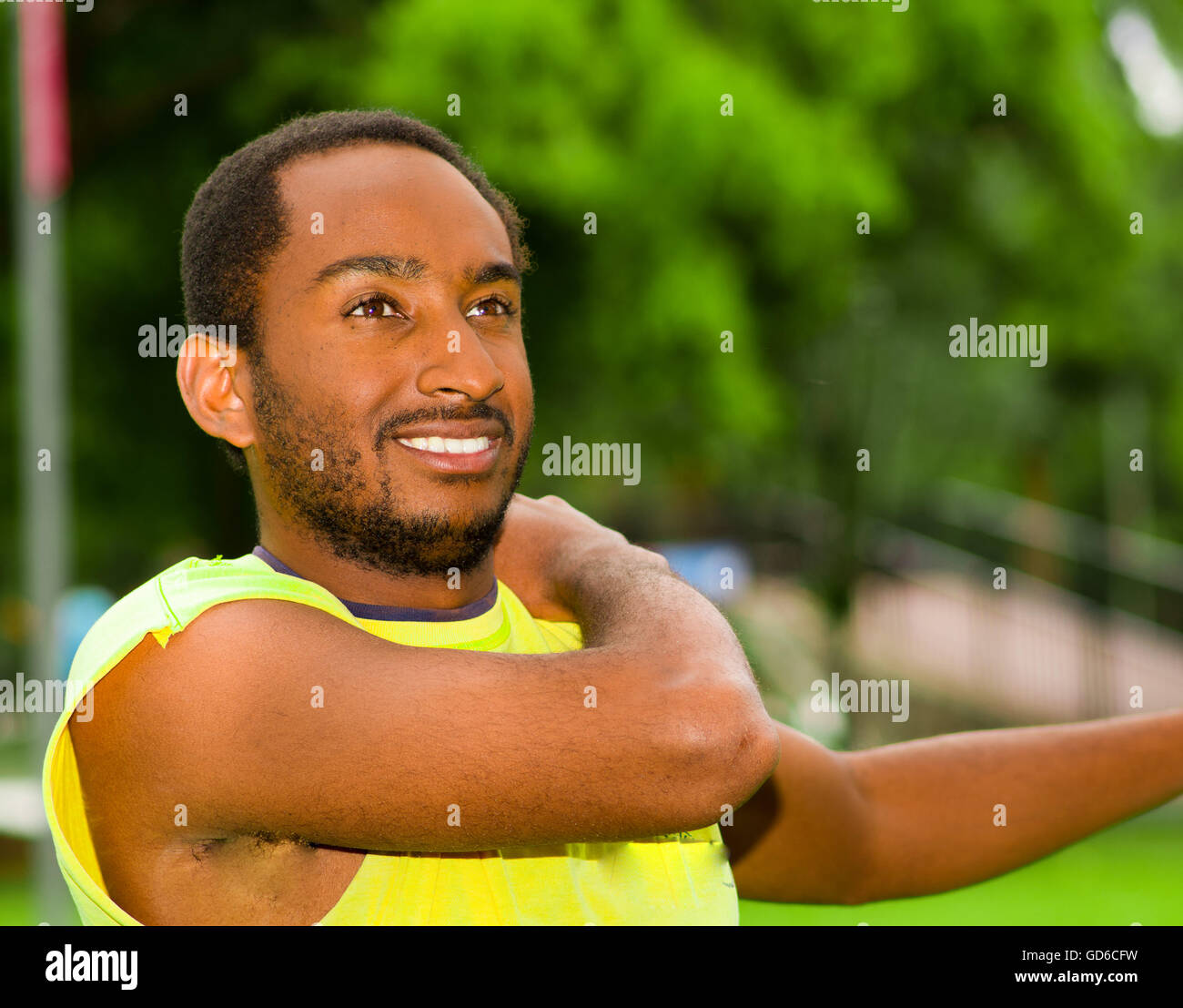 Man wearing yellow shirt stretching arms in park sorrounded by green ...