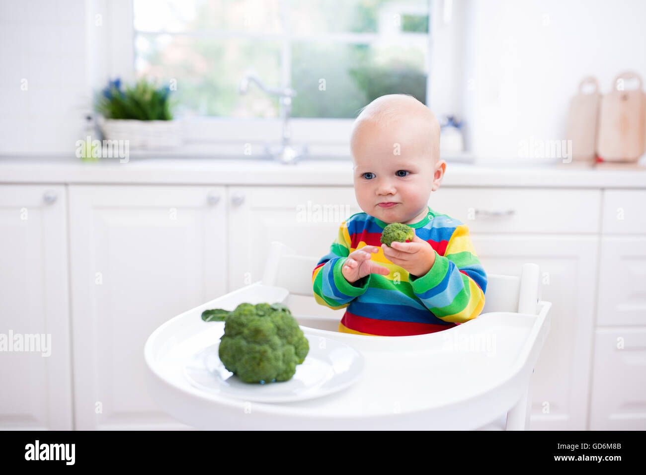 Happy baby sitting in high chair eating broccoli in a white kitchen ...