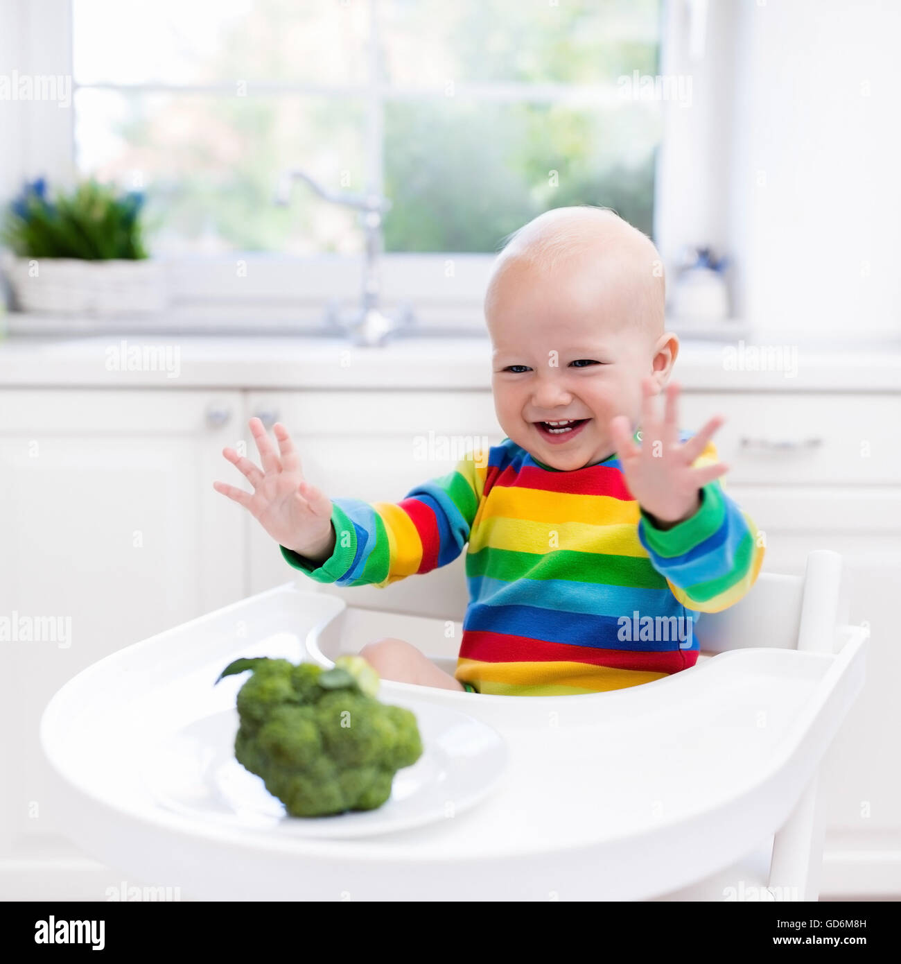 Happy baby sitting in high chair eating broccoli in a white kitchen ...