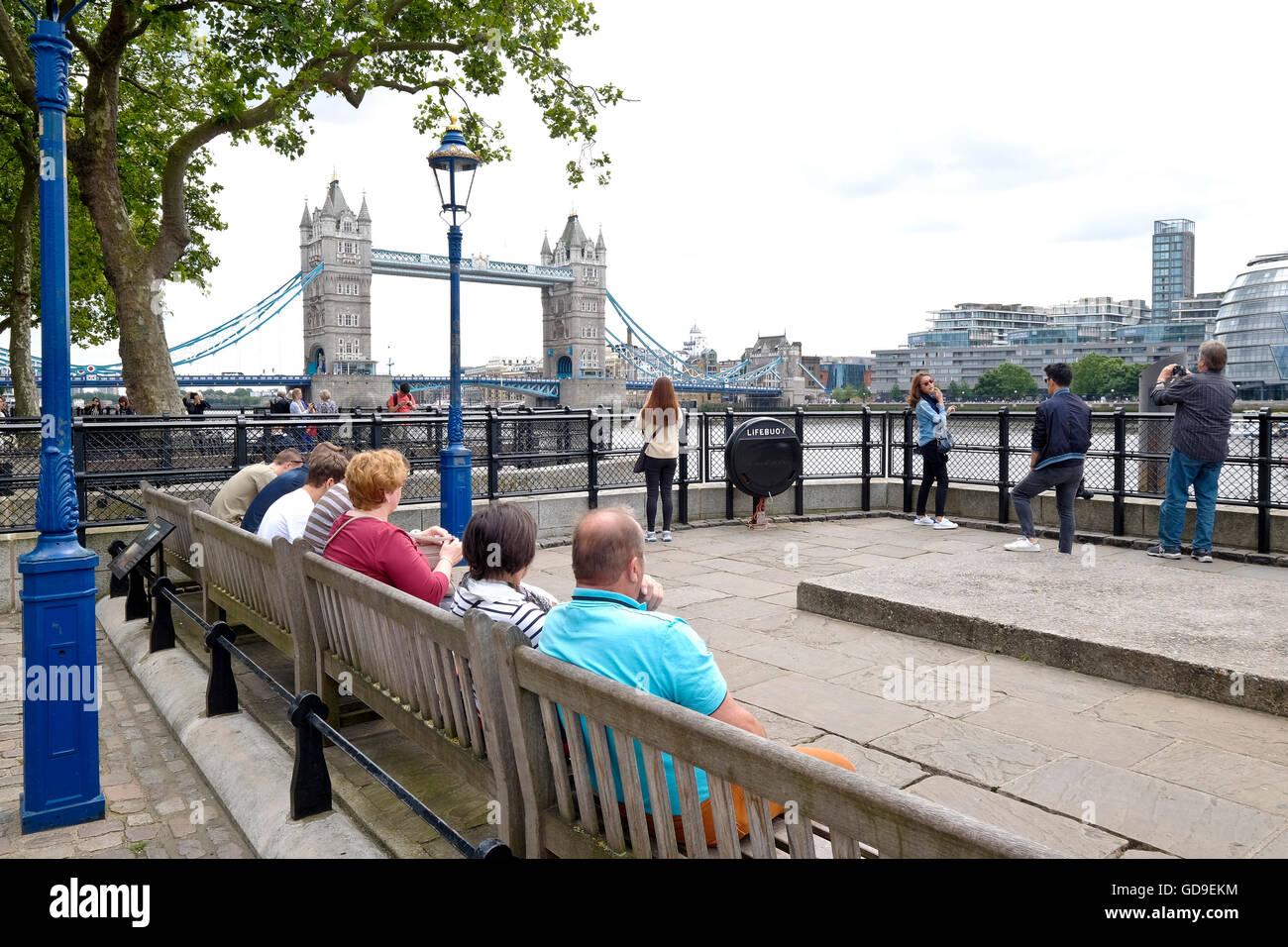 Tourists sit on benches Victoria Embankment alongside the Thames. Tower ...