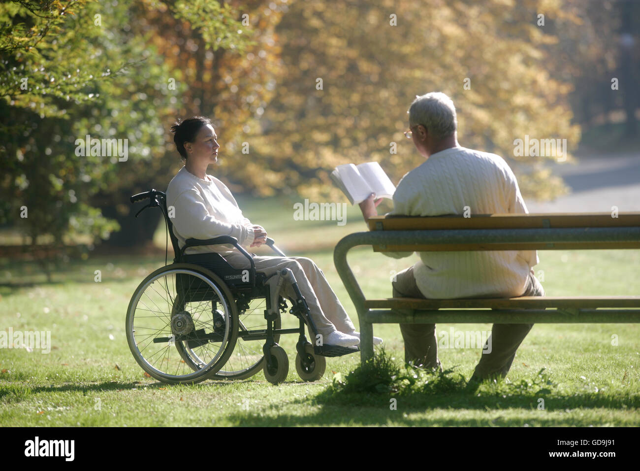 Woman sitting in a wheelchair, man sitting on a park bench with a book ...