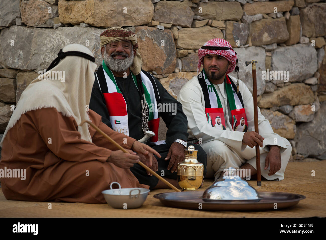 Three Emirati men in traditional dress sit talking on straw mats in ...