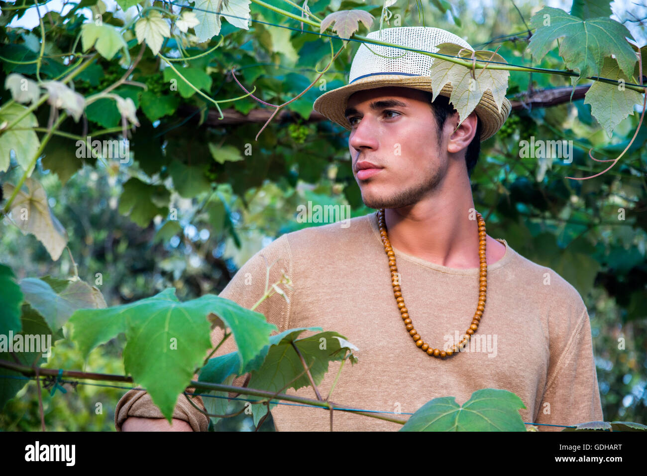 Side view of handsome young man in hat toching vine leaves in garden in ...