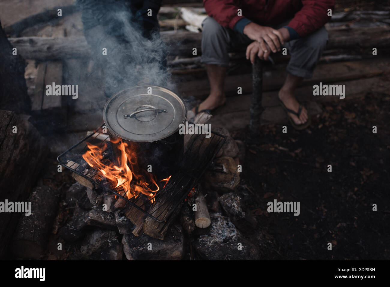 Senior man sitting on log, watching cooking pot on campfire, low ...