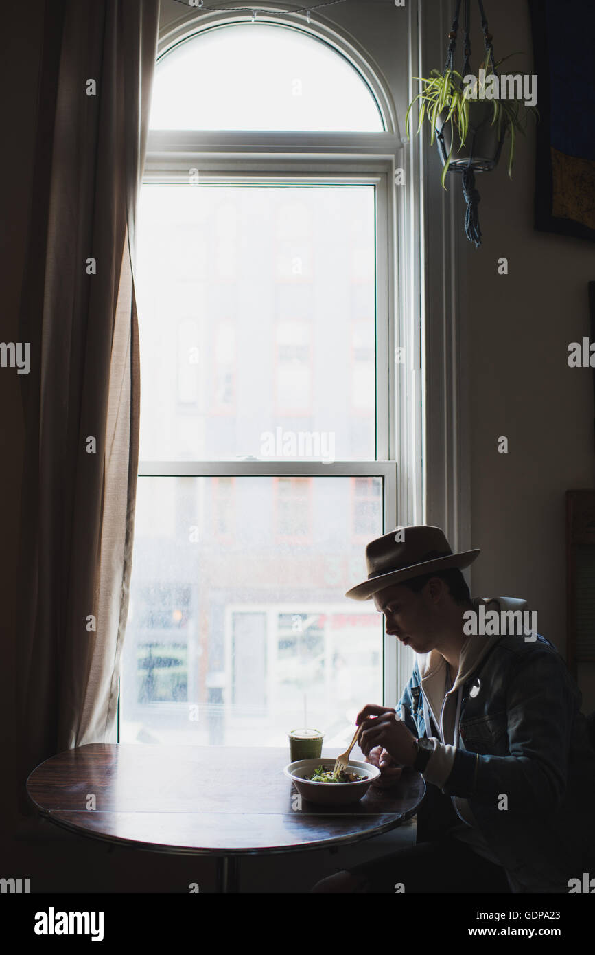 Young man sitting at table by window, eating healthy meal Stock Photo ...