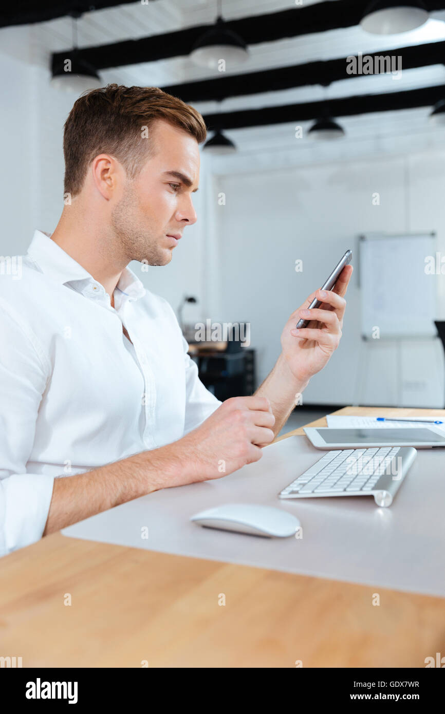 Serious young businessman sitting at workplace and using cell phone ...