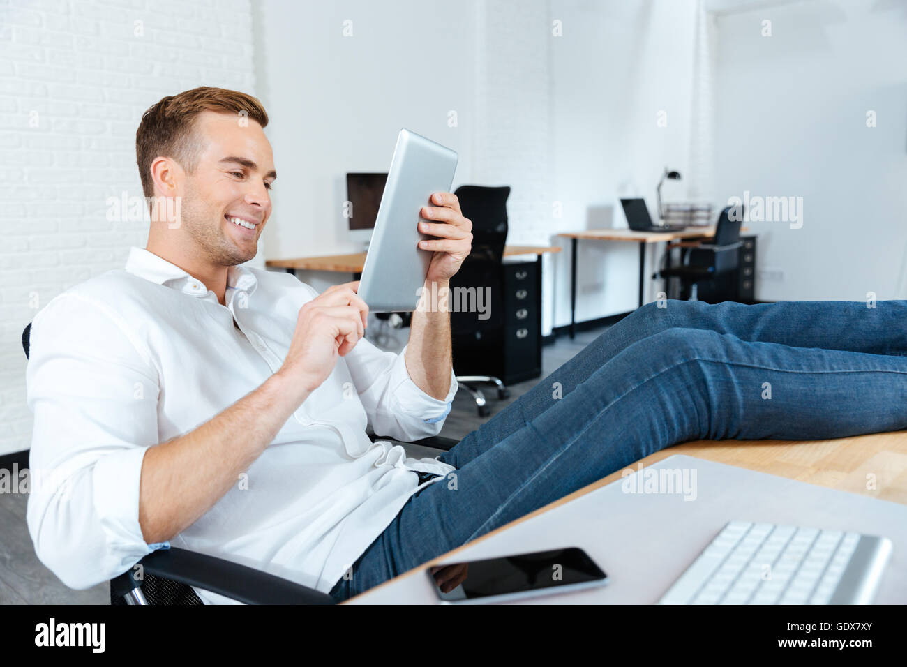 Happy young businessman sitting with legs on table and using tablet at ...
