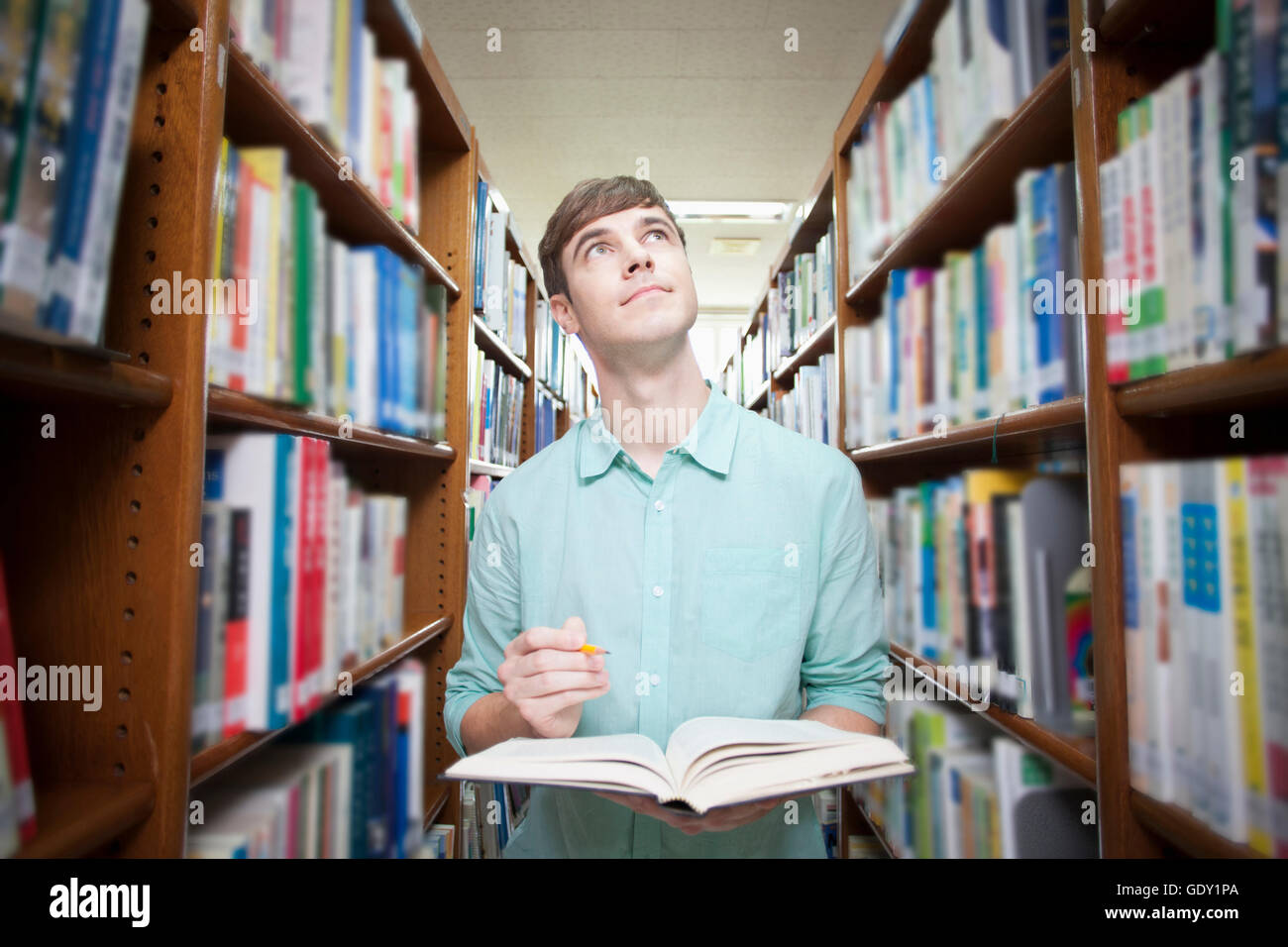 Portrait of young man holding a book and a pencil looking up at library ...