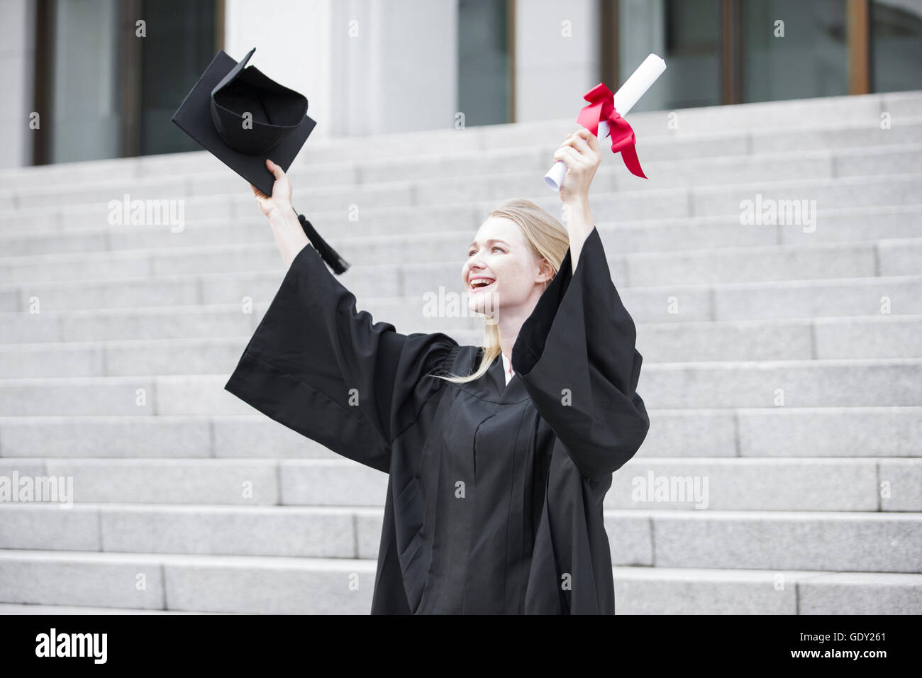 Smiling college graduate in gown holding her hat and diploma looking up ...