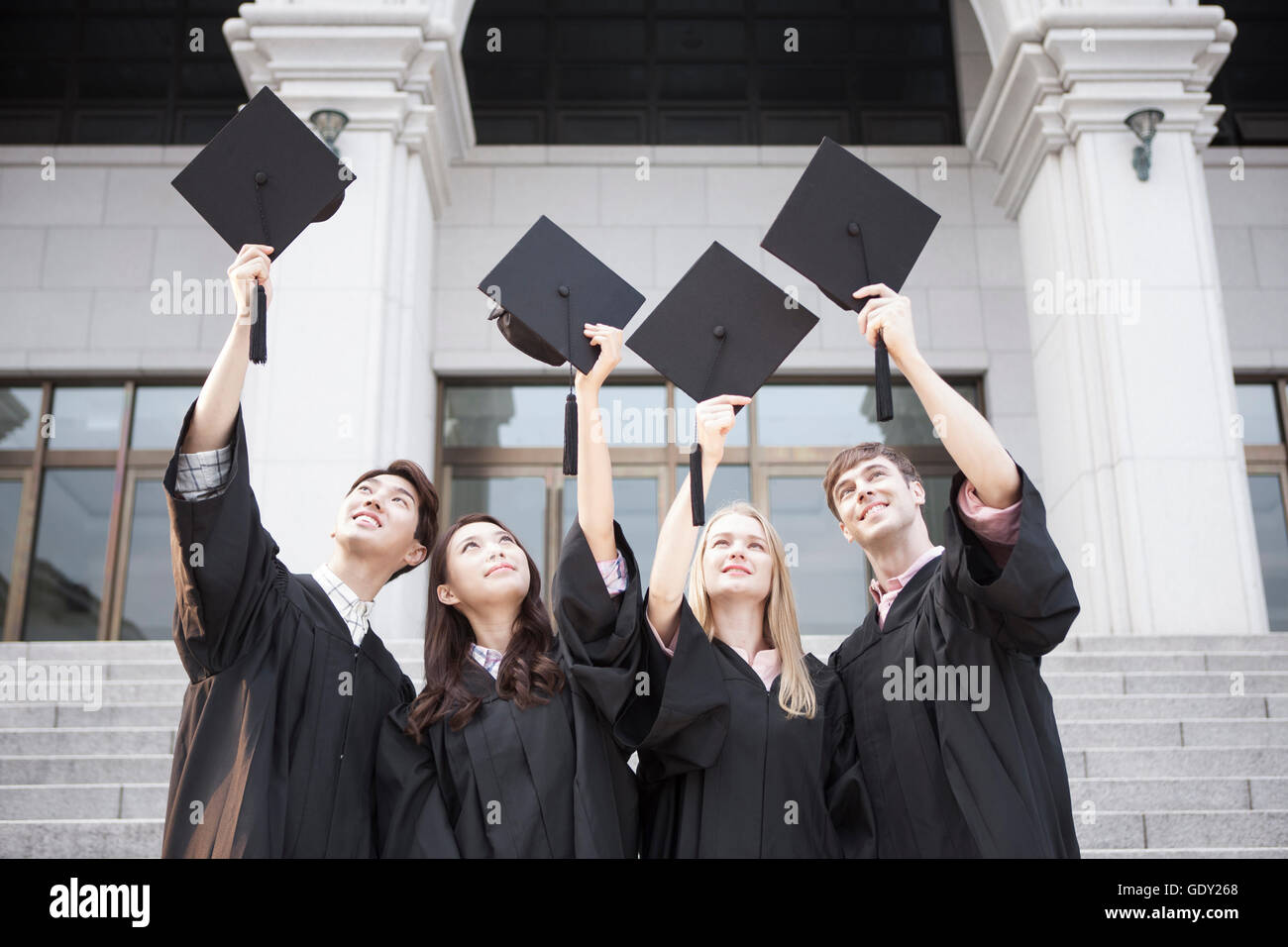 Portrait of smiling college graduates in gowns holding hats looking up ...