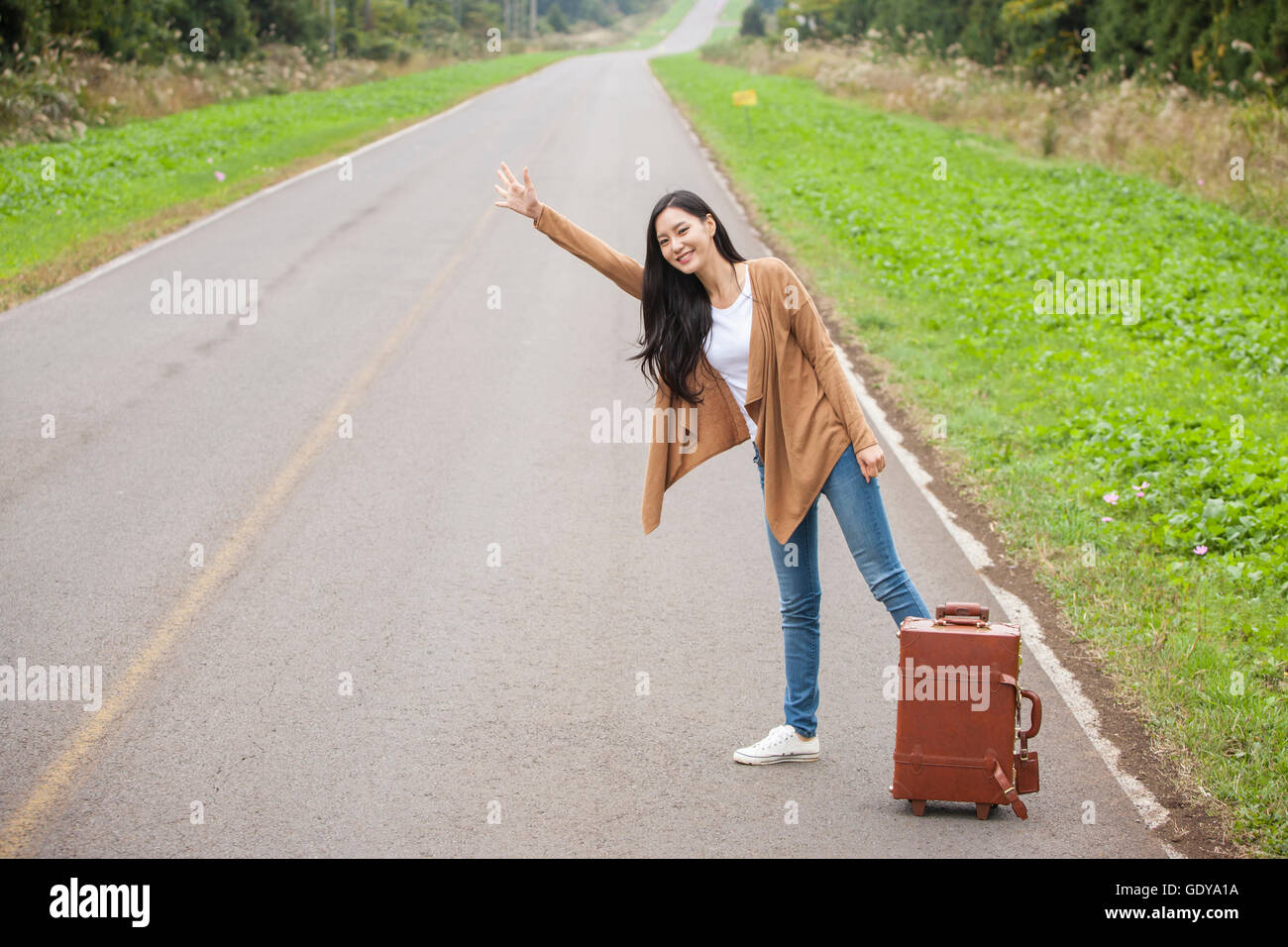 Young smiling female hitchhiker standing raising her hand on roadside ...
