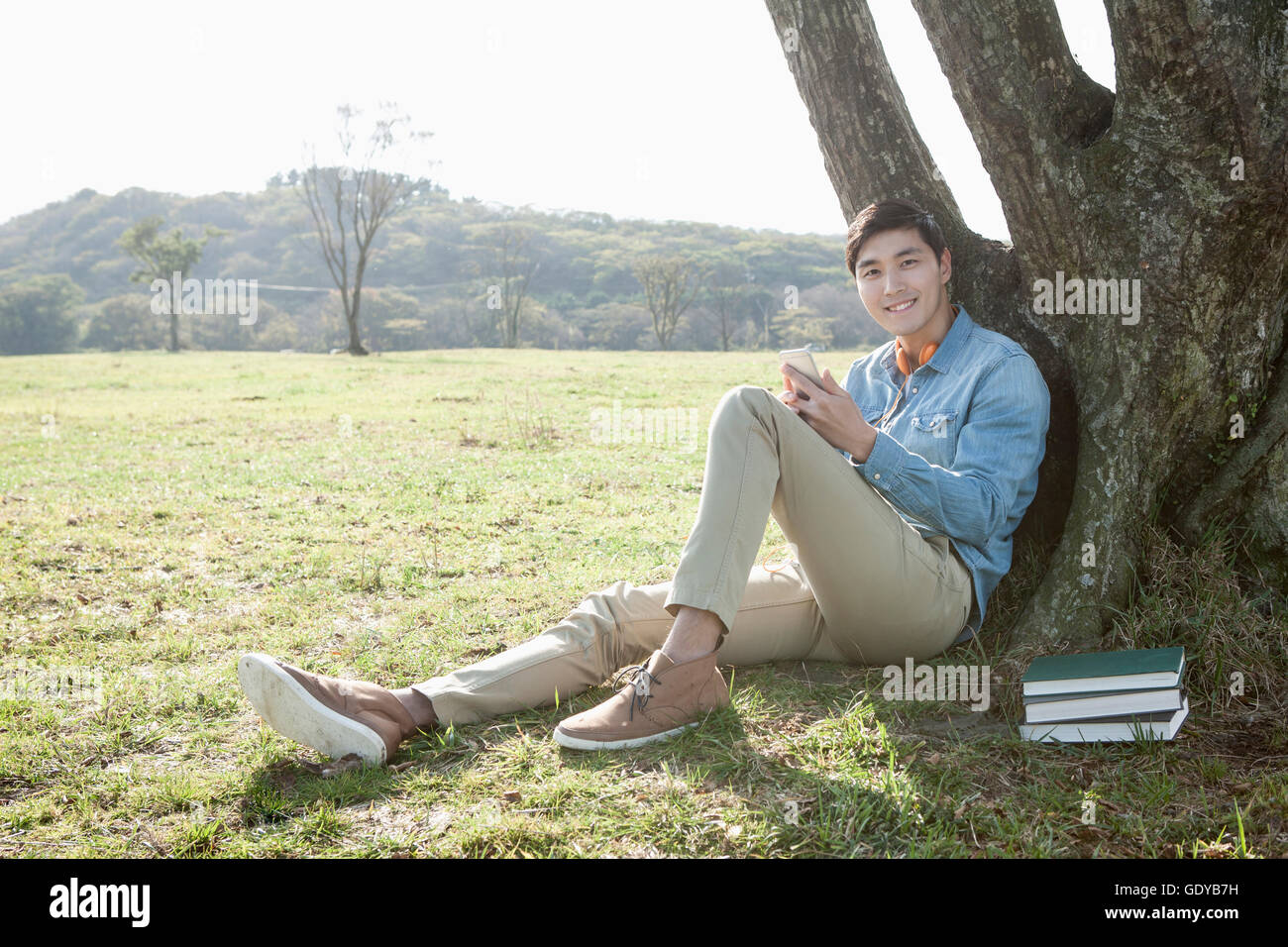 Young smiling man sitting and resting under a tree on grassland staring ...