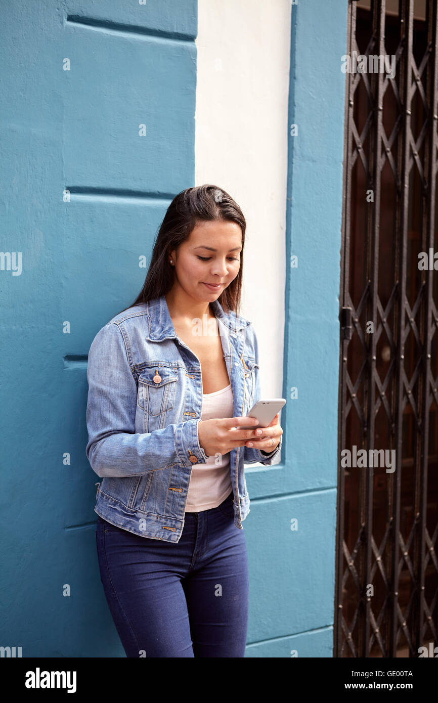 Pretty young girl typing on her cell phone while leaning against a blue ...