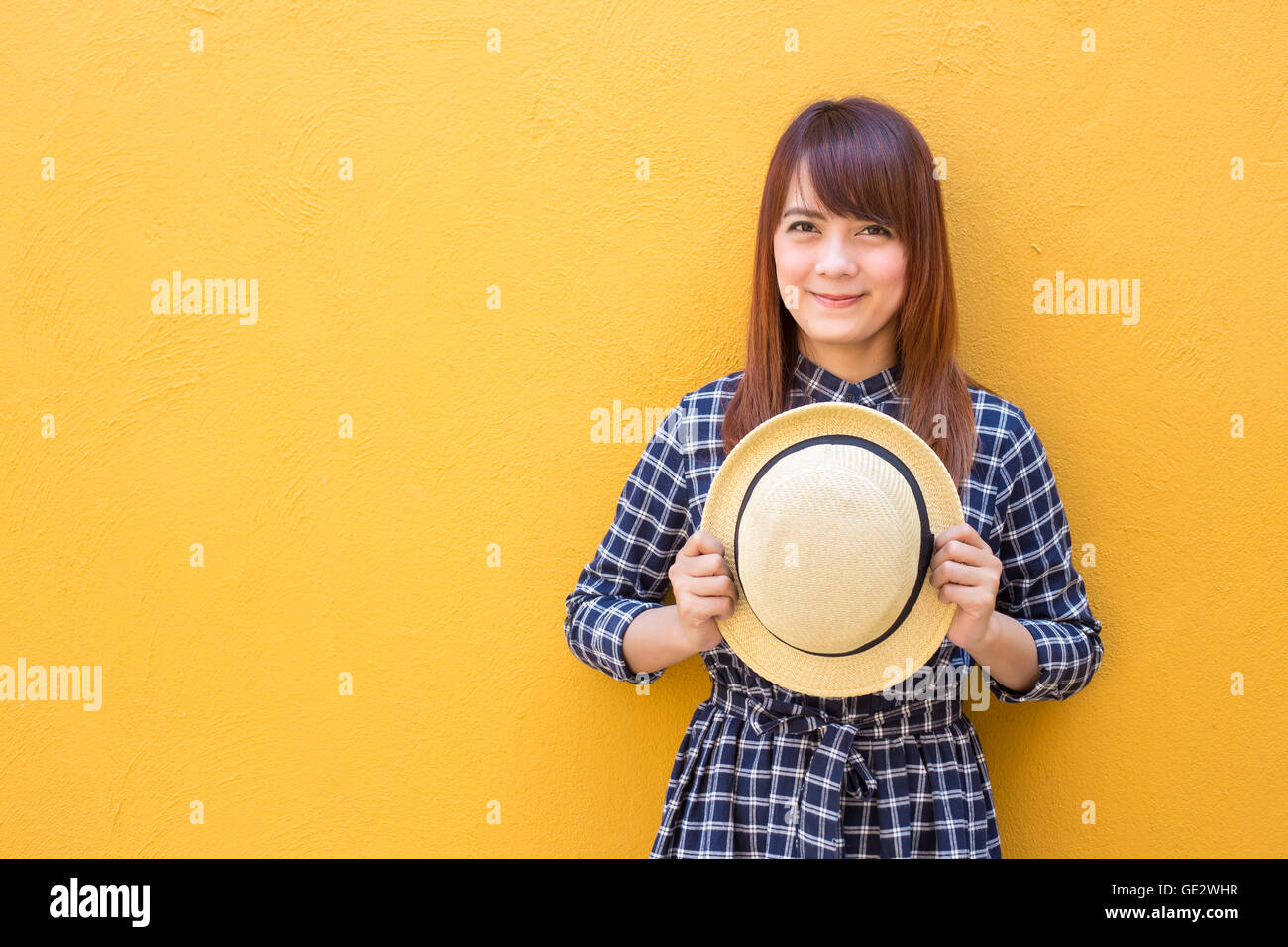 smiling woman wear in dress hand holding hat on yellow cement wall ...
