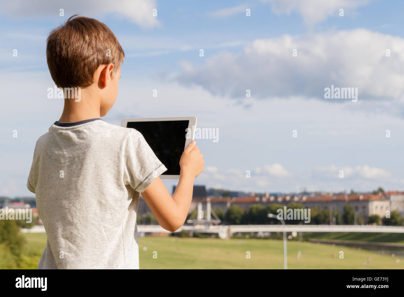 Smiling boy hold tablet PC. Outdoor. Blue sky and city background. Back ...