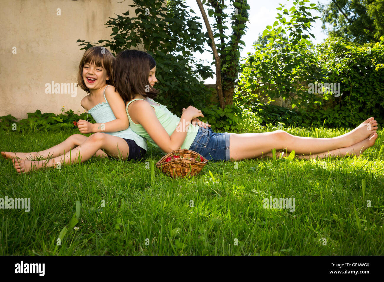 Two laughing sisters sitting back to back on a meadow Stock Photo - Alamy