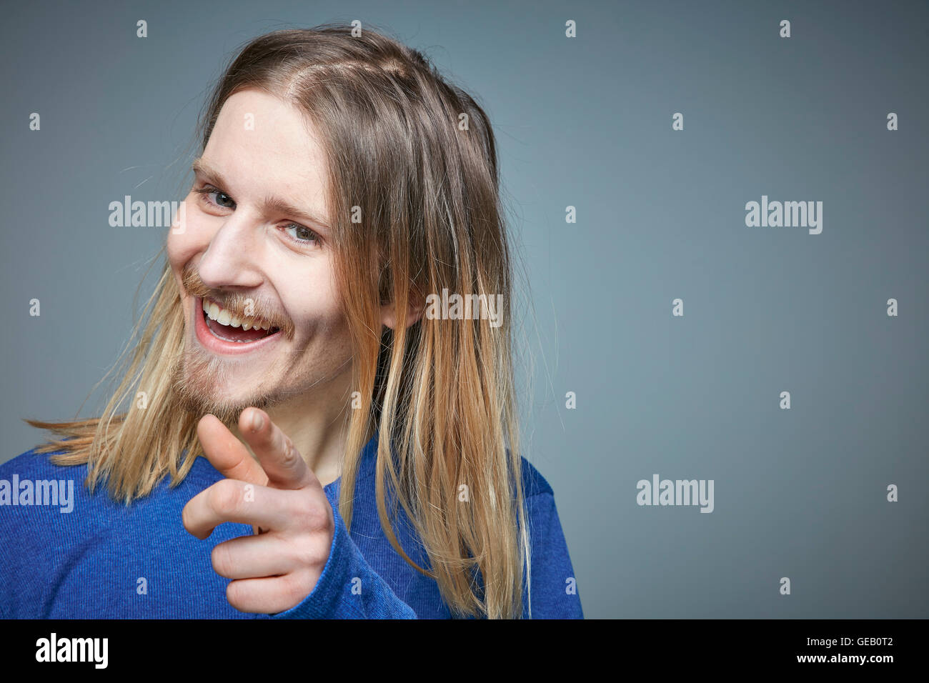 Portrait of laughing young man with long blond hair pointing on viewer ...