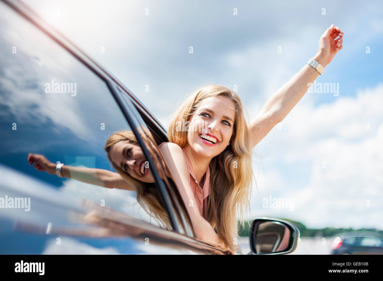 Portrait of smiling young woman leaning out of car window raising arm ...