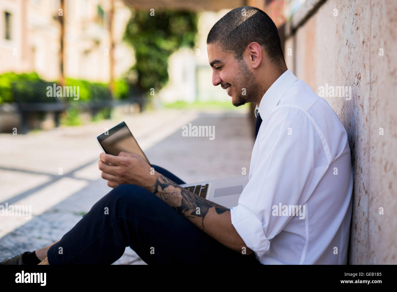 Smiling young businessman sitting on the ground looking at his tablet ...