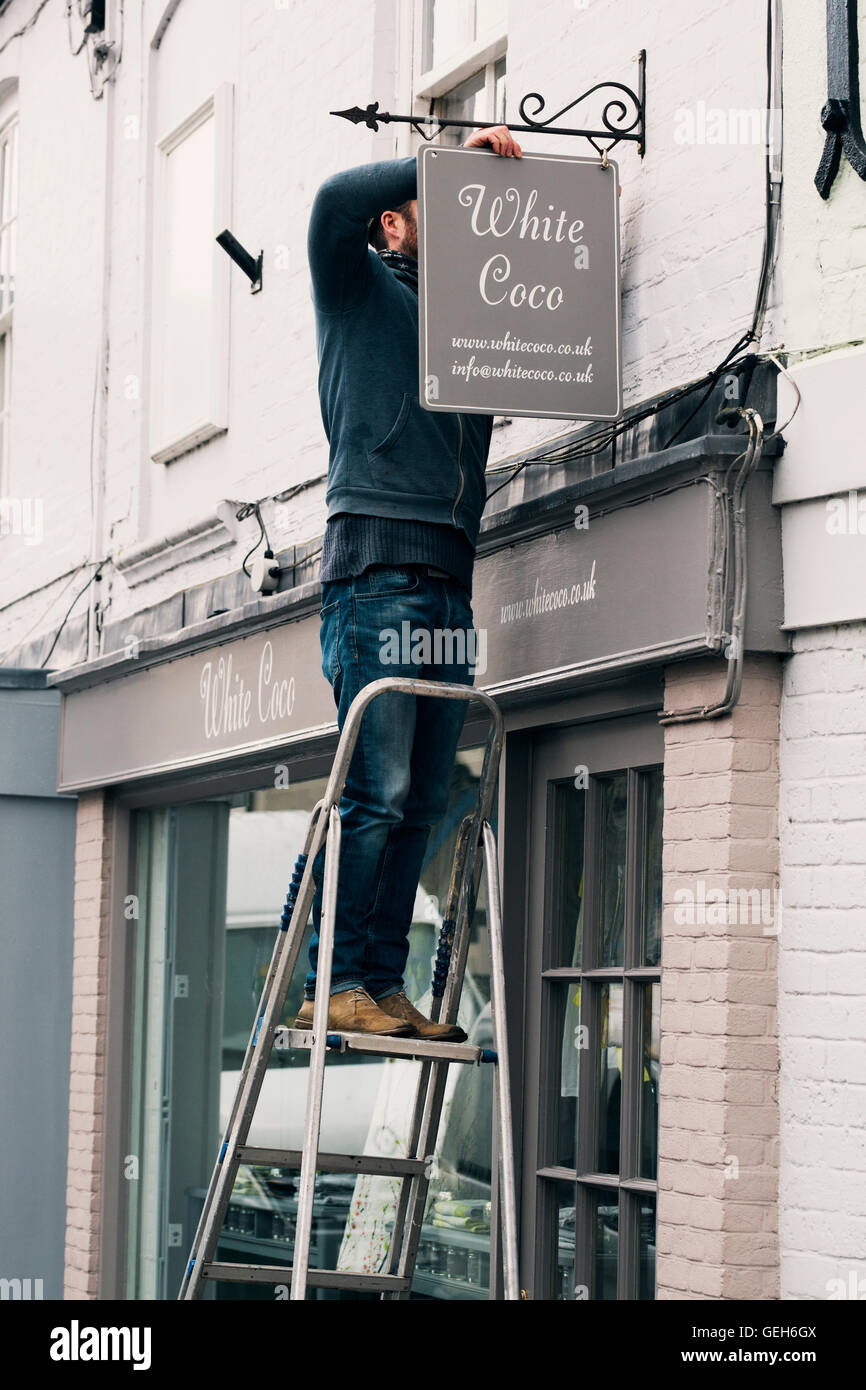 A man on a ladder fixing a painted name sign onto a bracket on a ...
