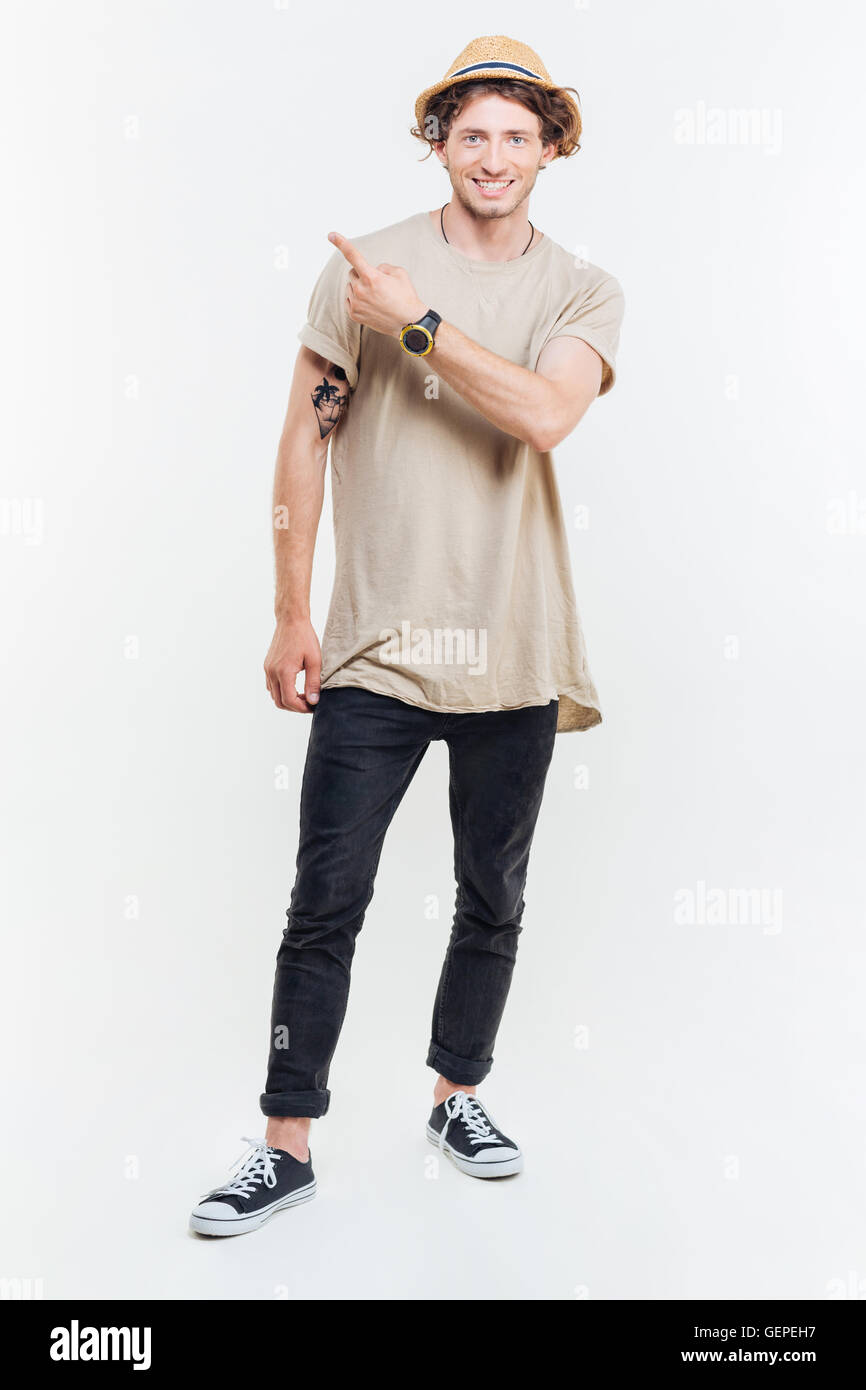 Smiling young man in a hat standing and pointing away over white ...
