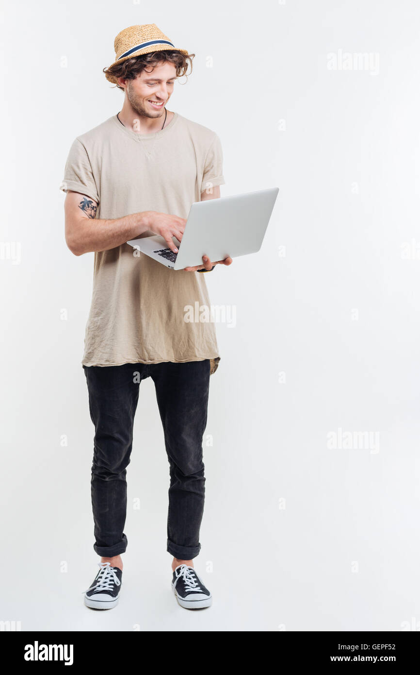 Cheerful young man in hat standing and using laptop over white ...
