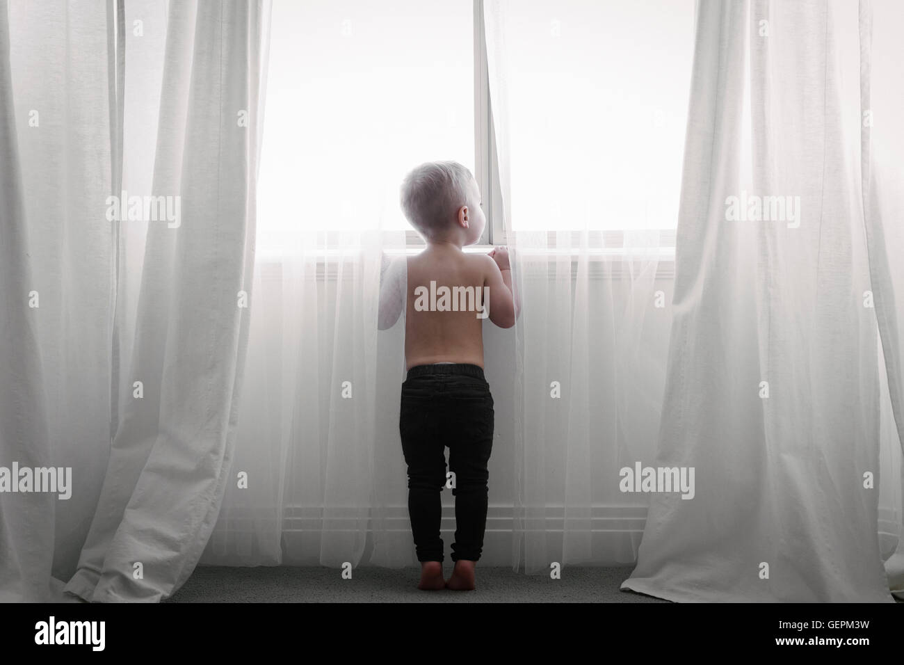 A child standing at a window looking out through the net curtains. Back ...