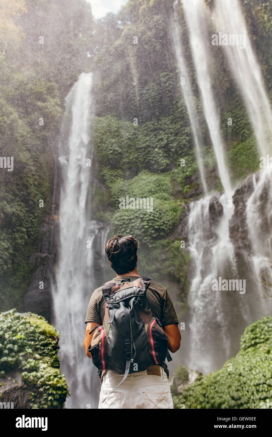 Rear view of young man with backpack standing in front of waterfall ...