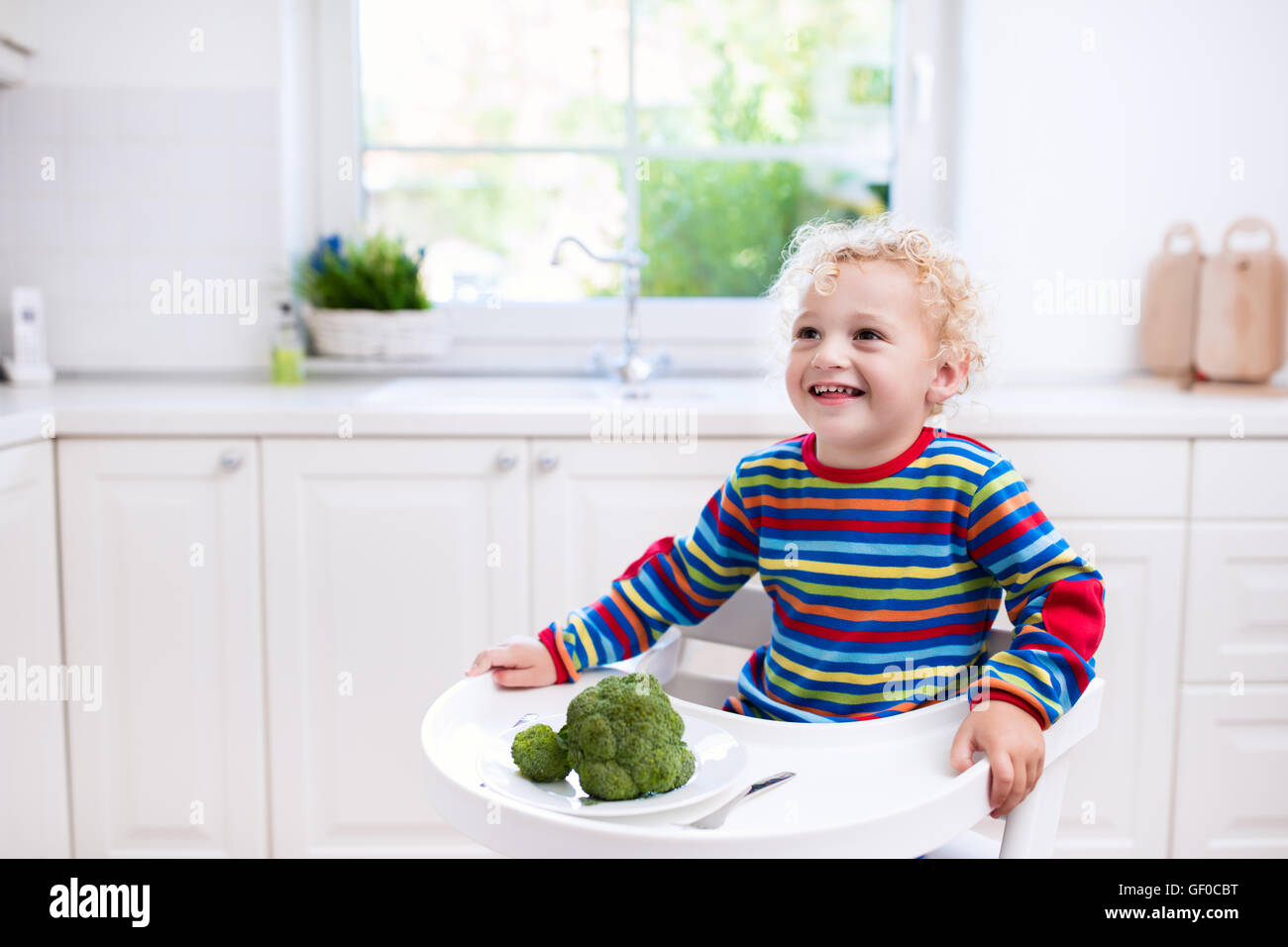 Happy baby sitting in high chair eating broccoli in a white kitchen ...