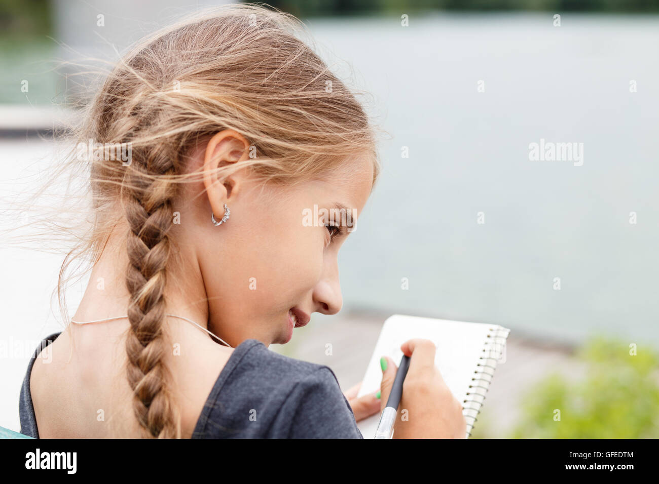 Young girl with braids drawing sketch in notebook sitting near a pond ...