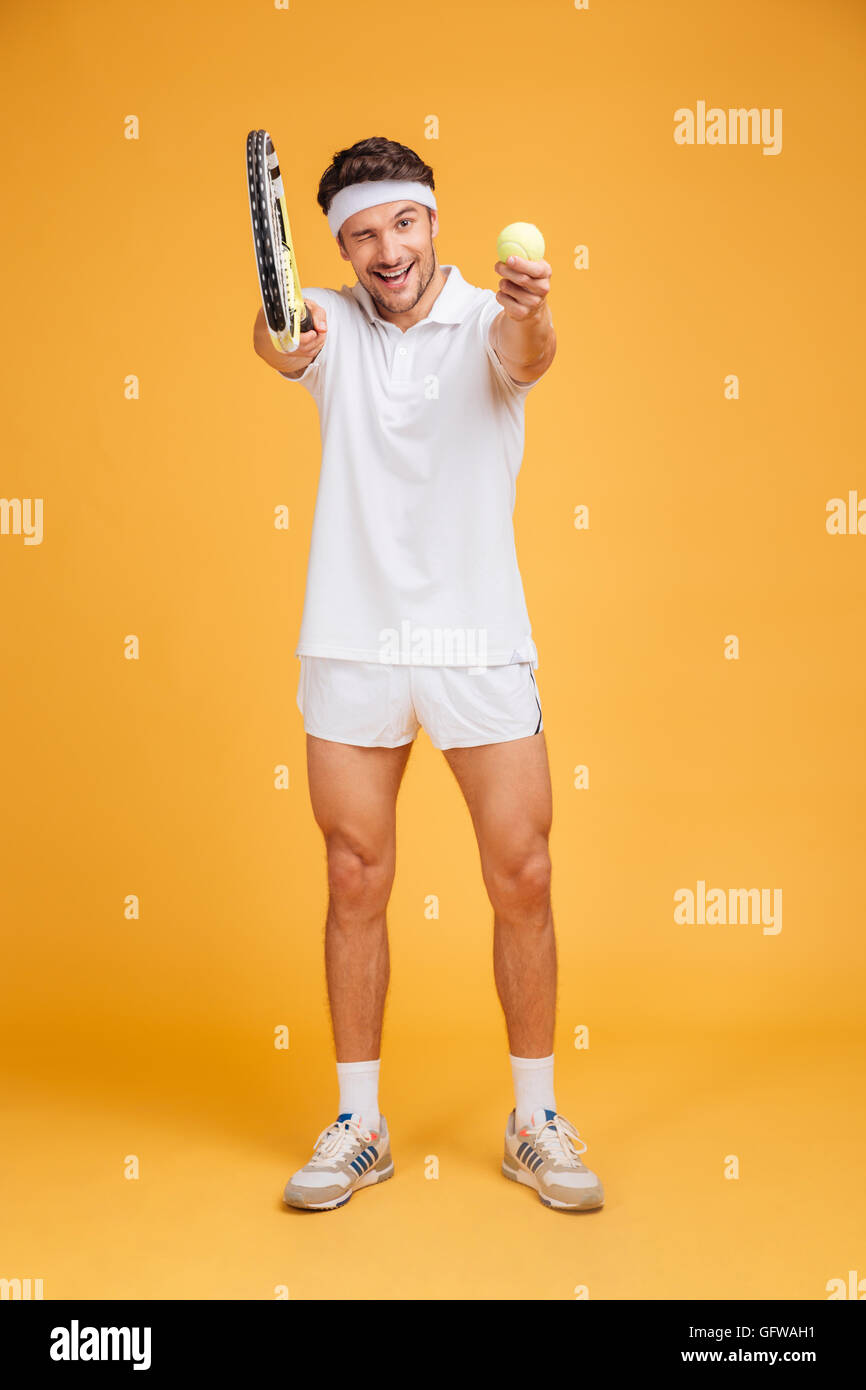 Cheerful playful young man tennis player winking and giving you ball ...