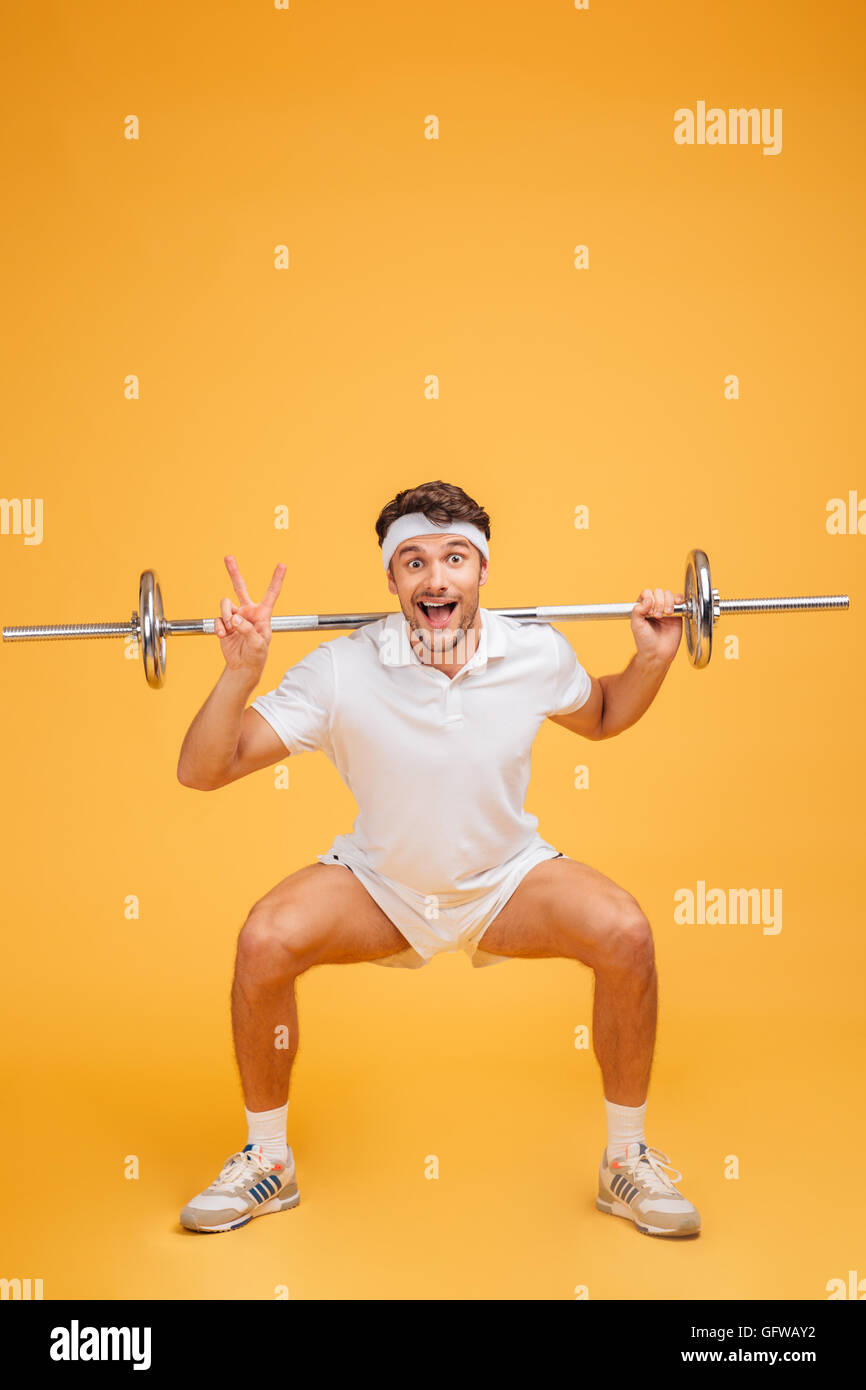 Funny cheerful young sportsman doing squats with barbell and showing ...
