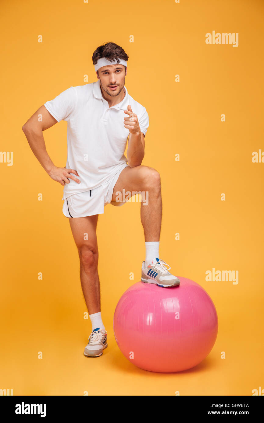 Young handsome sportsman with one foot on fitness ball and pointing at ...