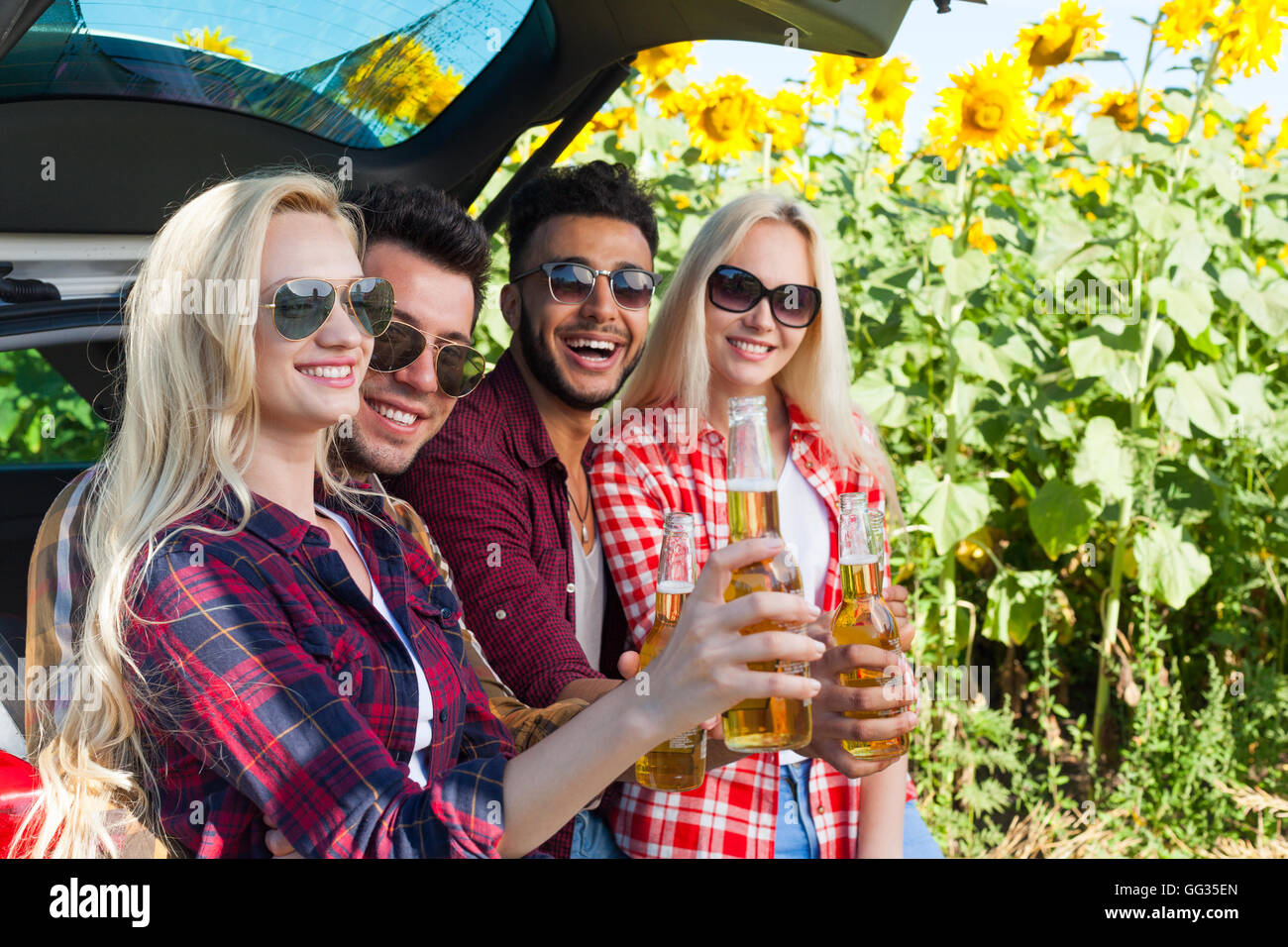 Friends drinking beer toasting clink bottles sitting in car trunk ...