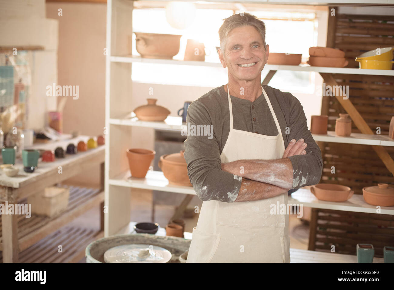 Smiling male potter standing with arms crossed in pottery workshop ...